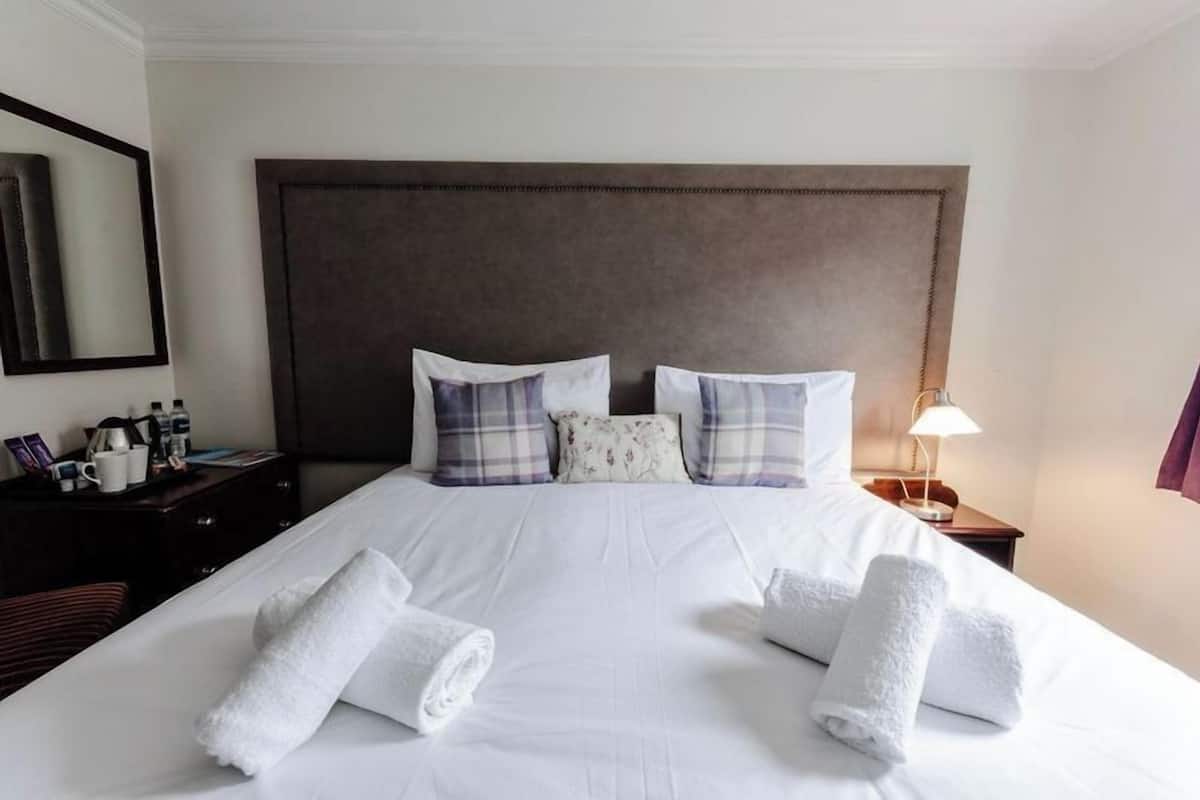 Cama bem arrumada com lençóis brancos e duas toalhas enroladas, cabeceira escura e mesa de cabeceira com abajur.