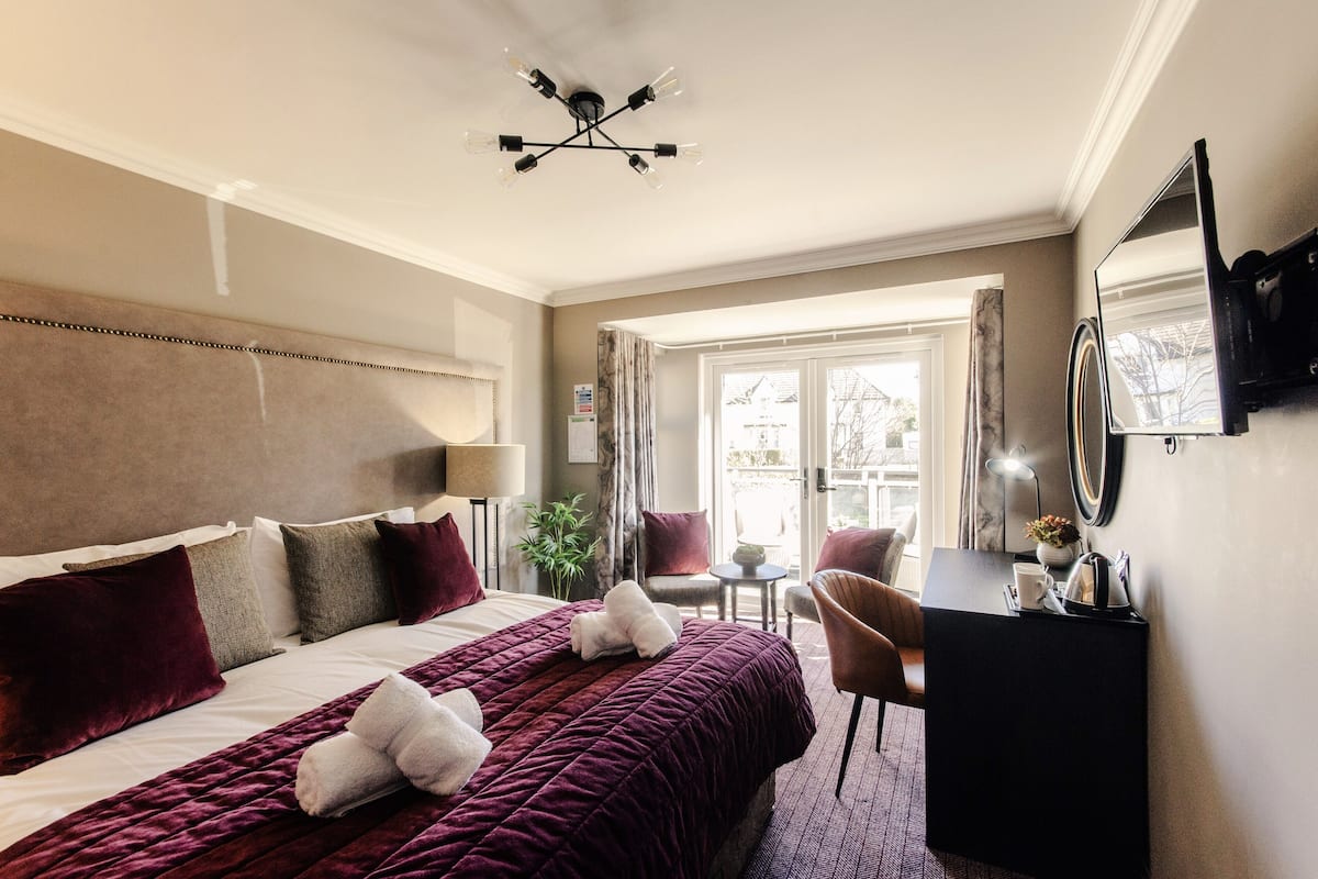 Quarto de hotel com uma cama grande, uma escrivaninha, uma cadeira, uma televisão e uma janela com cortinas.