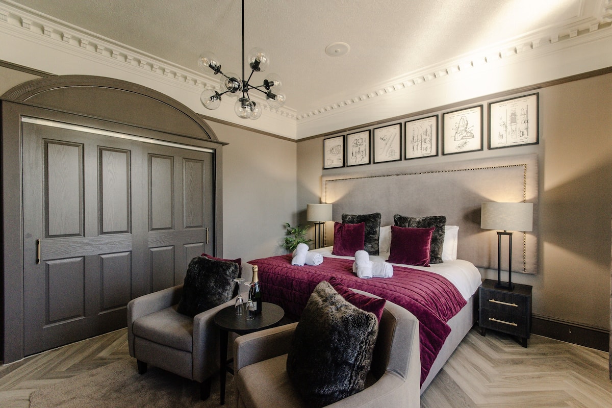 Quarto de hotel com uma cama grande, duas poltronas, uma mesinha, um criado-mudo e um candelabro.