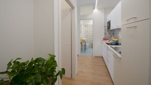 Um corredor estreito que dá para um banheiro e uma área de cozinha, com armários brancos e piso de madeira.