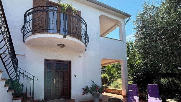 Um edifício de dois andares com varanda, área de estar externa com cadeiras de vime e uma mesa, e um vaso de planta.