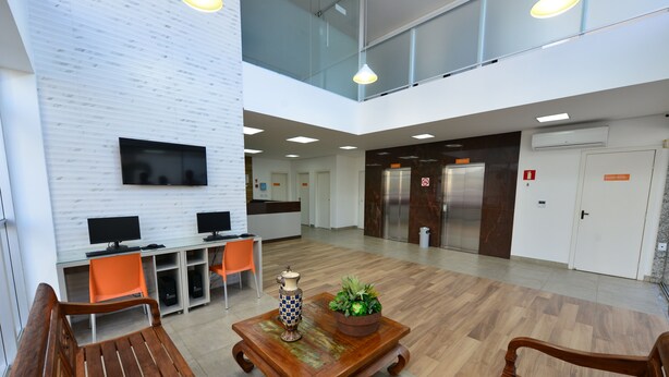 Um hall amplo com teto alto, balcão de recepção, duas estações de computador, mesa de centro com vaso decorativo e planta, e um banco de madeira.