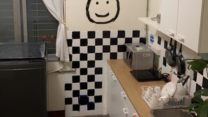 Uma cozinha compacta com painel decorativo xadrez, uma decoração com carinha sorridente, fogão embutido, pia e um pequeno espaço de refeições.