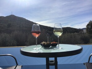 Uma mesa com duas taças de vinho e uma tigela de petiscos, com uma paisagem campestre ao fundo.