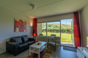 Sala de estar com um sofá preto, uma mesa de centro branca e uma cadeira. Há uma janela grande com vista para uma paisagem verde.