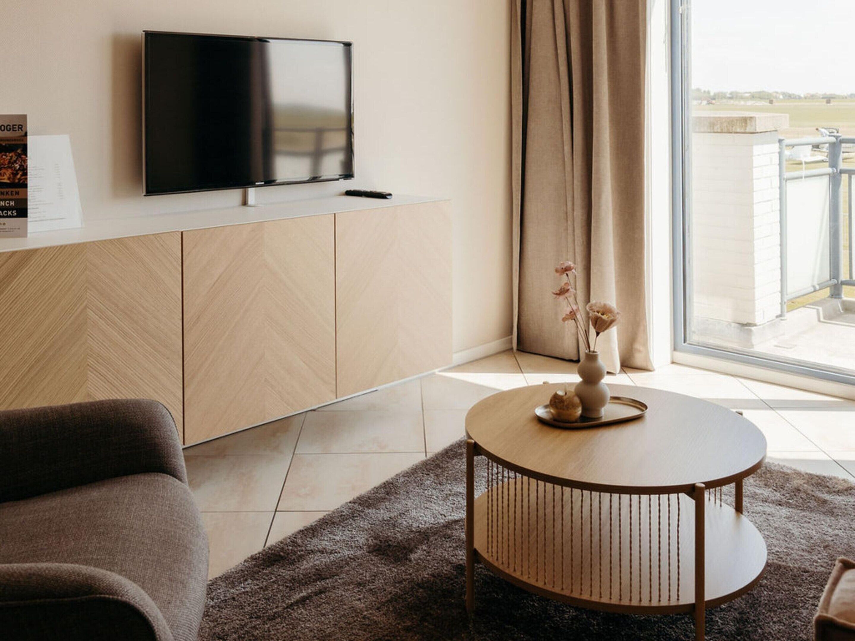 Uma sala de estar moderna com uma TV de tela plana presa a um armário de madeira clara, uma mesa de centro redonda com um vaso de flores secas e vista para uma varanda através de janelas amplas.