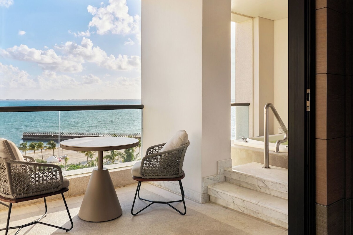 Uma varanda com vista para o mar, uma mesa redonda, duas cadeiras de vime e uma escadaria que dá para um espaço interno.