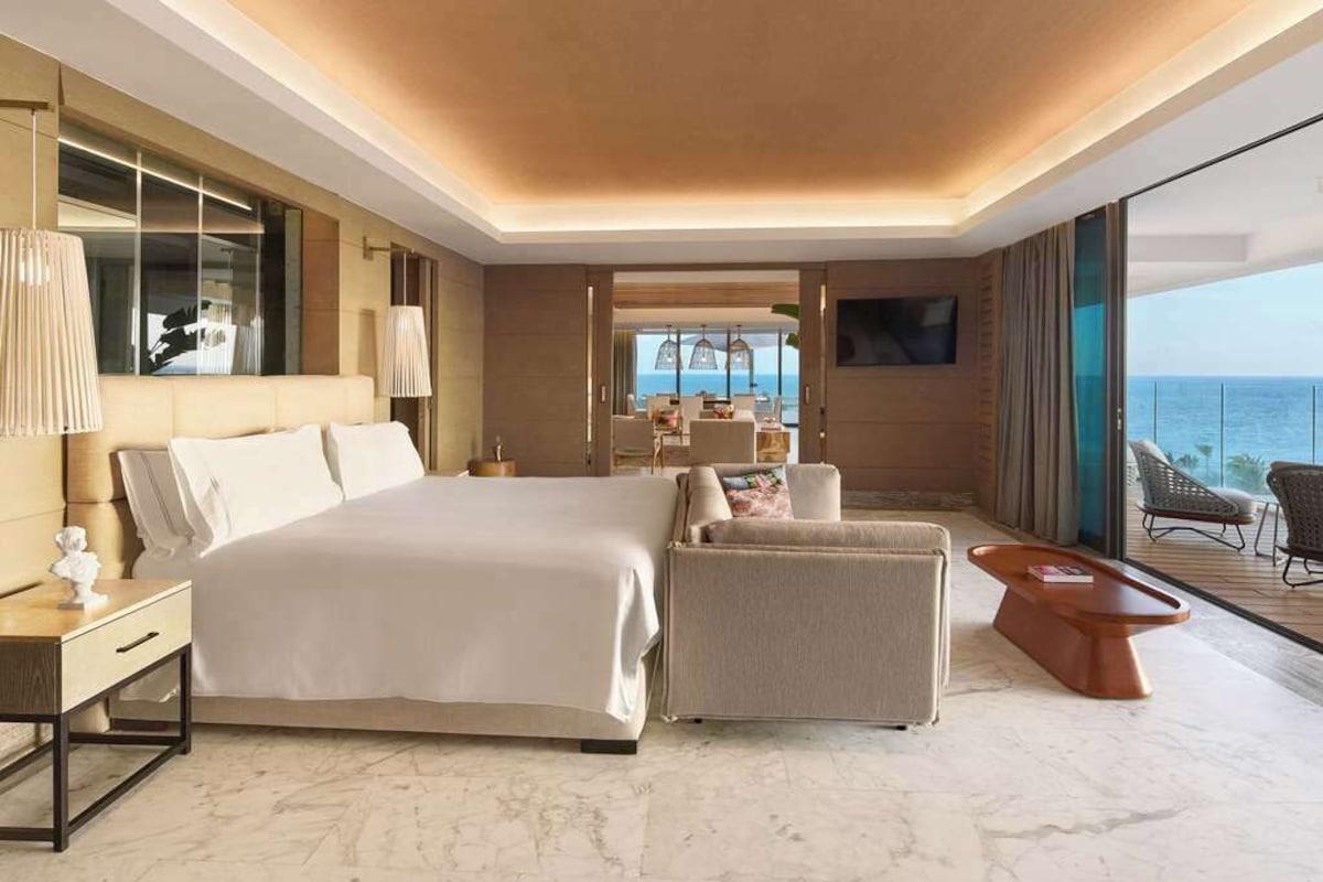 Quarto de hotel moderno com uma cama grande, área de estar com vista para o oceano e varanda com mobiliário externo.