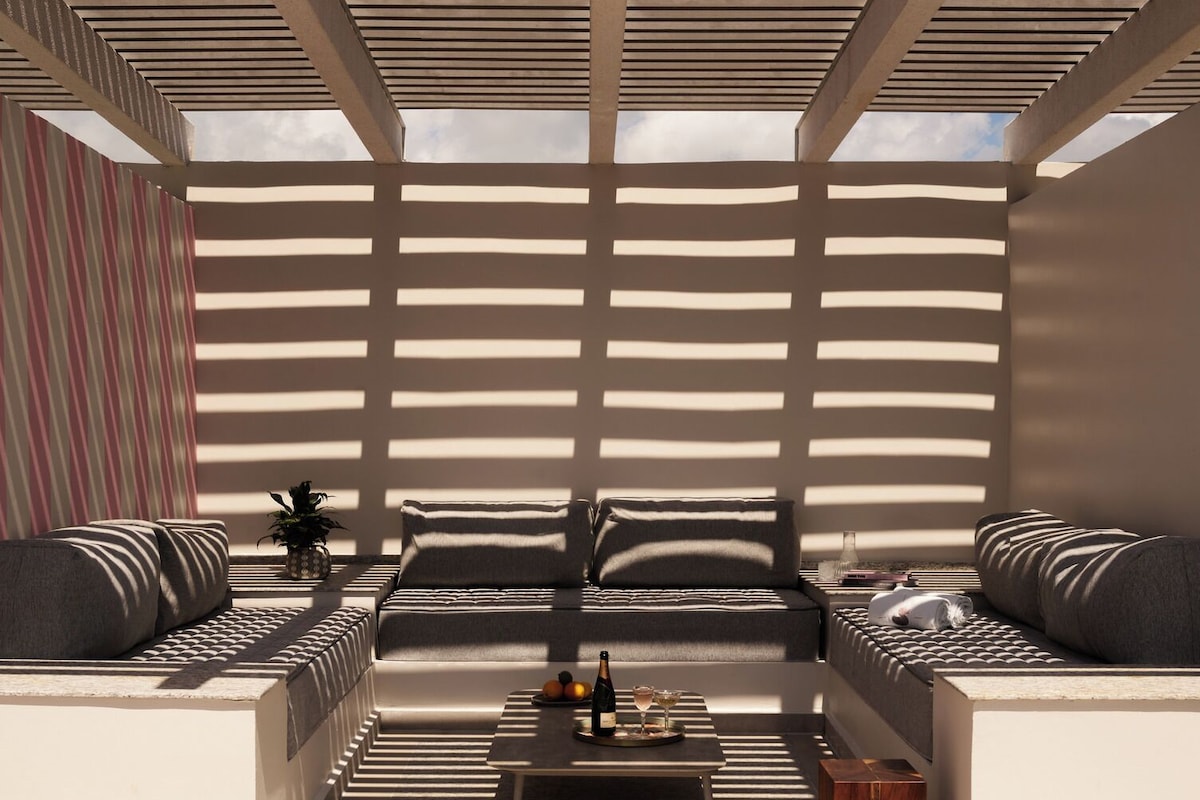 Uma área externa moderna com assentos, incluindo um sofá e duas poltronas, uma mesa pequena com bebidas e uma planta decorativa.