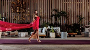 Uma mulher de vestido vermelho caminhando em um palco com um fundo que inclui um painel de madeira vertical e as letras "SLS".