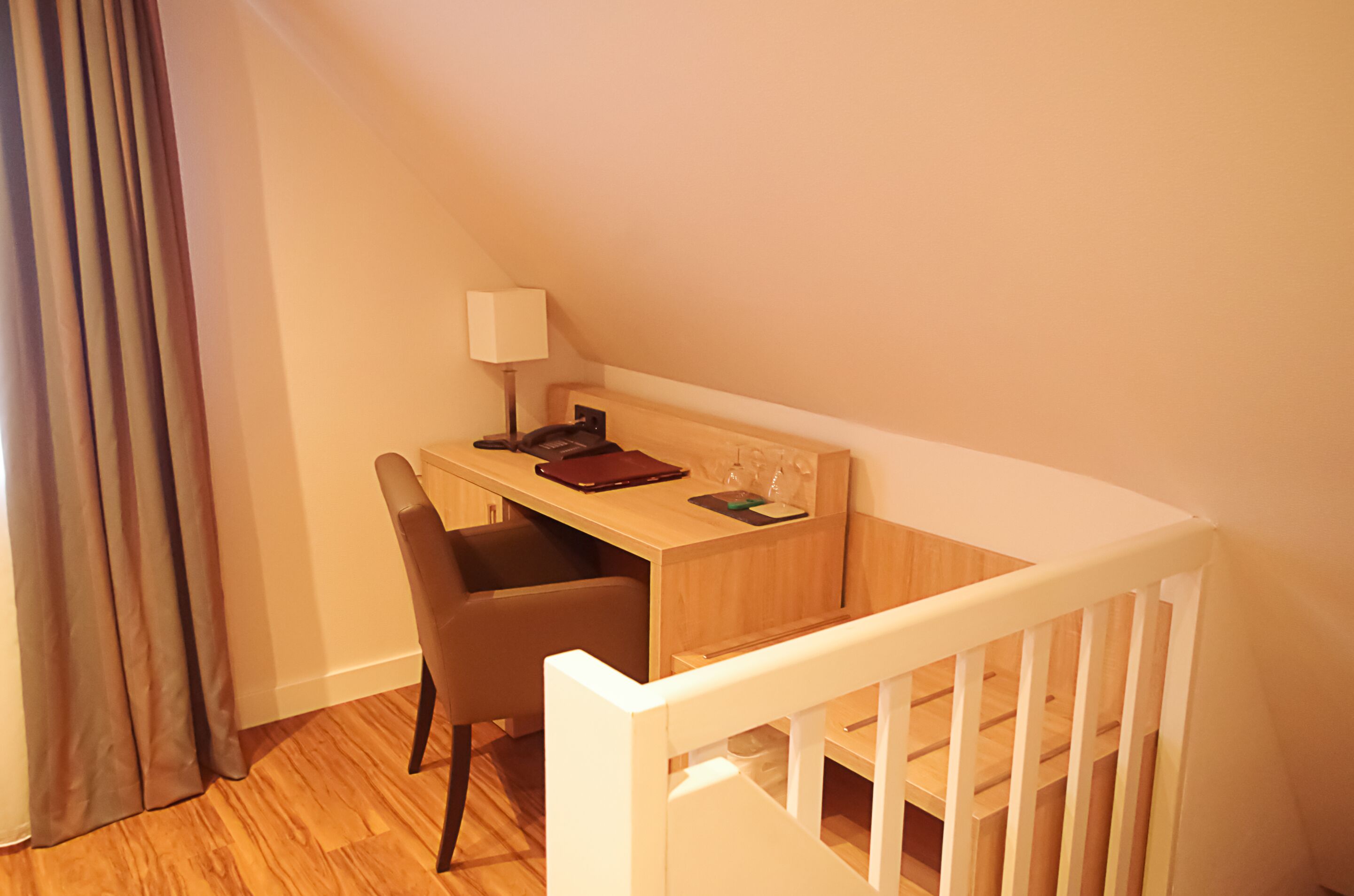 Quarto pequeno e bem iluminado, com escrivaninha de madeira, uma cadeira, abajur e berço branco.