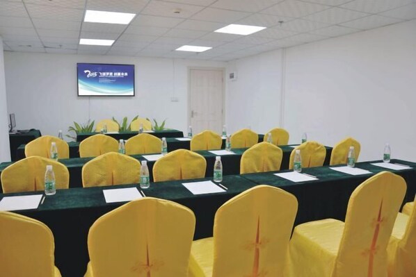 Uma sala de conferências com fileiras de cadeiras amarelas, uma mesa comprida com garrafas de água e uma tela exibindo texto.