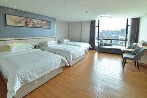 Quarto de hotel com duas camas, uma janela grande com vista para a cidade, área de refeições com mesa e cadeiras, e um quadro na parede.