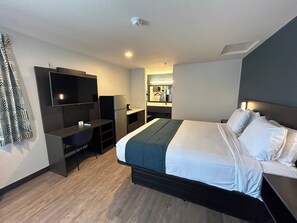 Quarto de hotel com cama, escrivaninha, televisão, espelho e cozinha compacta.