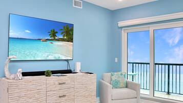 Quarto com uma TV grande exibindo uma cena de praia, um armário branco com gavetas, uma poltrona clara com almofada estampada e uma janela grande com varanda.