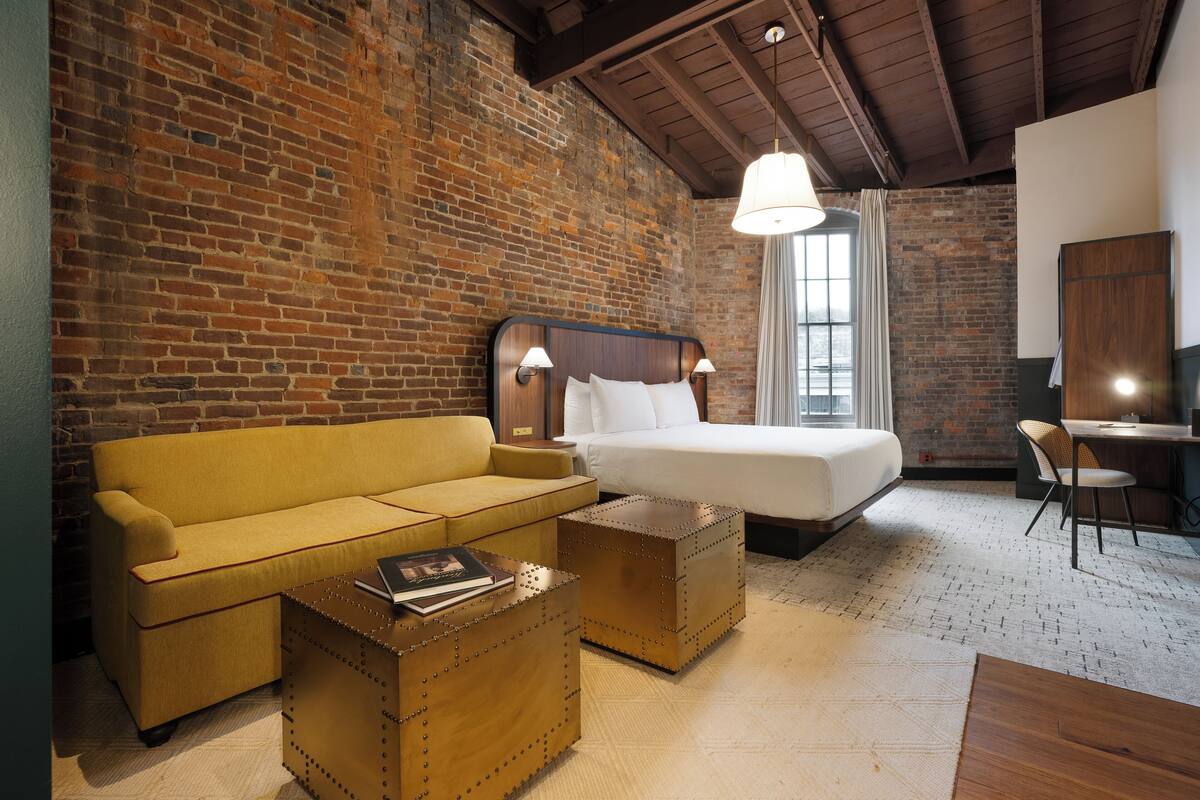 Quarto de hotel com parede de tijolos, sofá amarelo, cama com roupa de cama branca, mesa de centro de madeira e escrivaninha com cadeira.