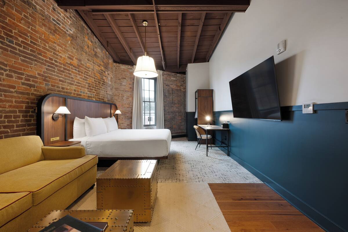 Quarto de hotel com parede de tijolos, cama, sofá, escrivaninha e televisão.
