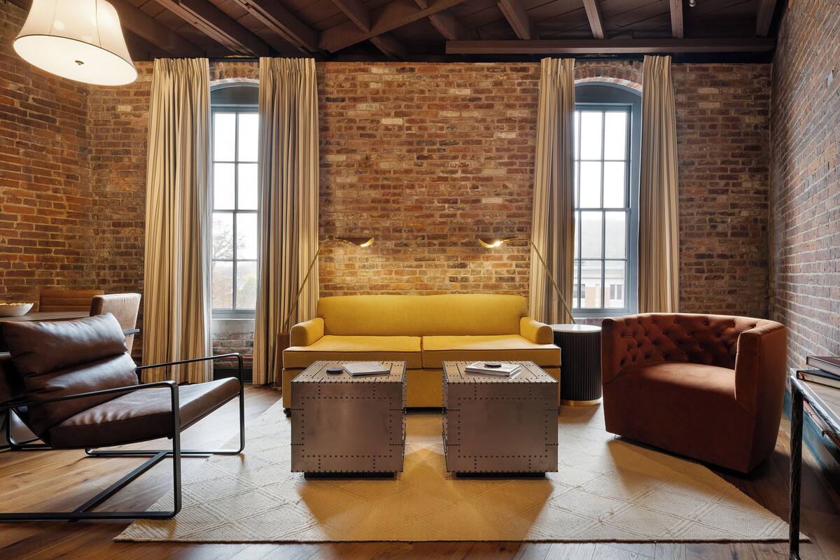 Uma sala de estar acolhedora com sofá amarelo, poltrona de couro e duas mesinhas de metal. Paredes de tijolo e grandes janelas com cortinas são visíveis.