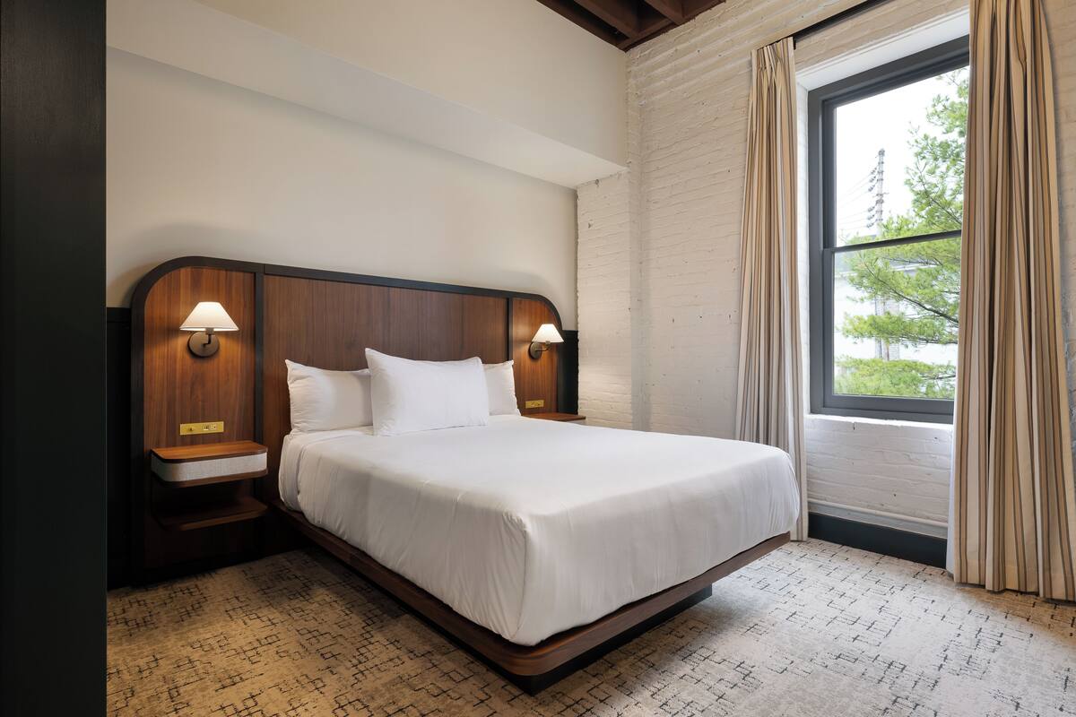 Cama bem arrumada com lençóis brancos, cabeceira de madeira, duas luminárias de cabeceira e uma janela com cortinas.