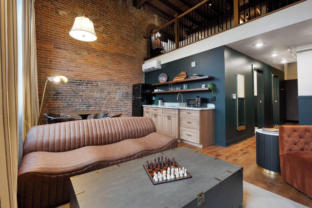 Um espaço de estar moderno em estilo loft, com cozinha no mezanino, uma ampla área de assentos acolchoados e um tabuleiro de xadrez sobre uma mesa de centro.