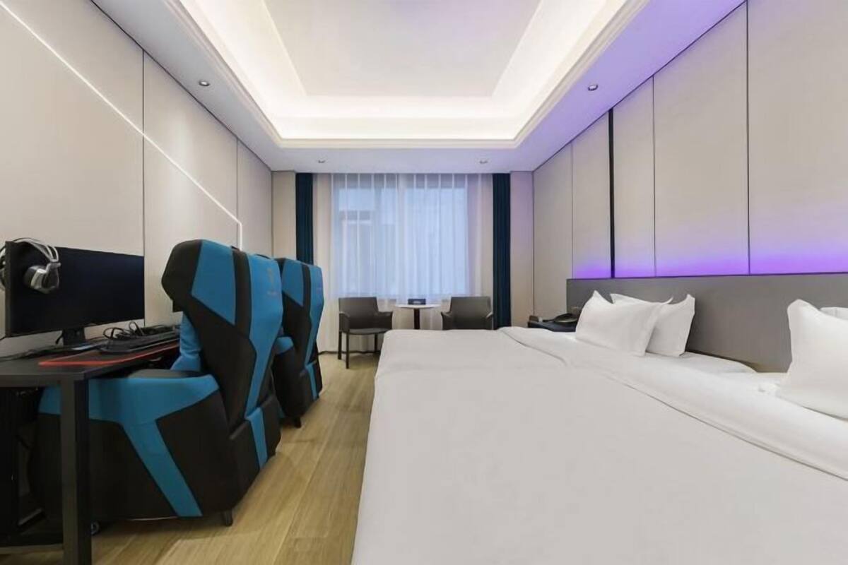 Quarto de hotel moderno com uma cama grande, uma escrivaninha com um computador, uma cadeira azul e preta e uma pequena mesa com uma cadeira.