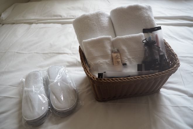 Cama com roupa branca, cesta de vime com produtos de higiene pessoal e um par de chinelos.
