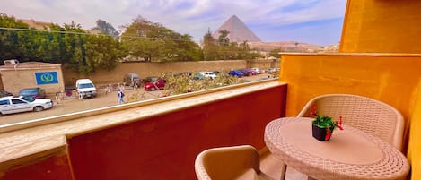 Varanda com cadeiras de vime e uma pequena mesa, com vista para um estacionamento e uma pirâmide ao longe.