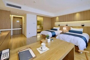 Quarto de hotel com duas camas, uma escrivaninha de madeira, uma mesinha com uma xícara e um livro, e uma mesa de cabeceira com um abajur.