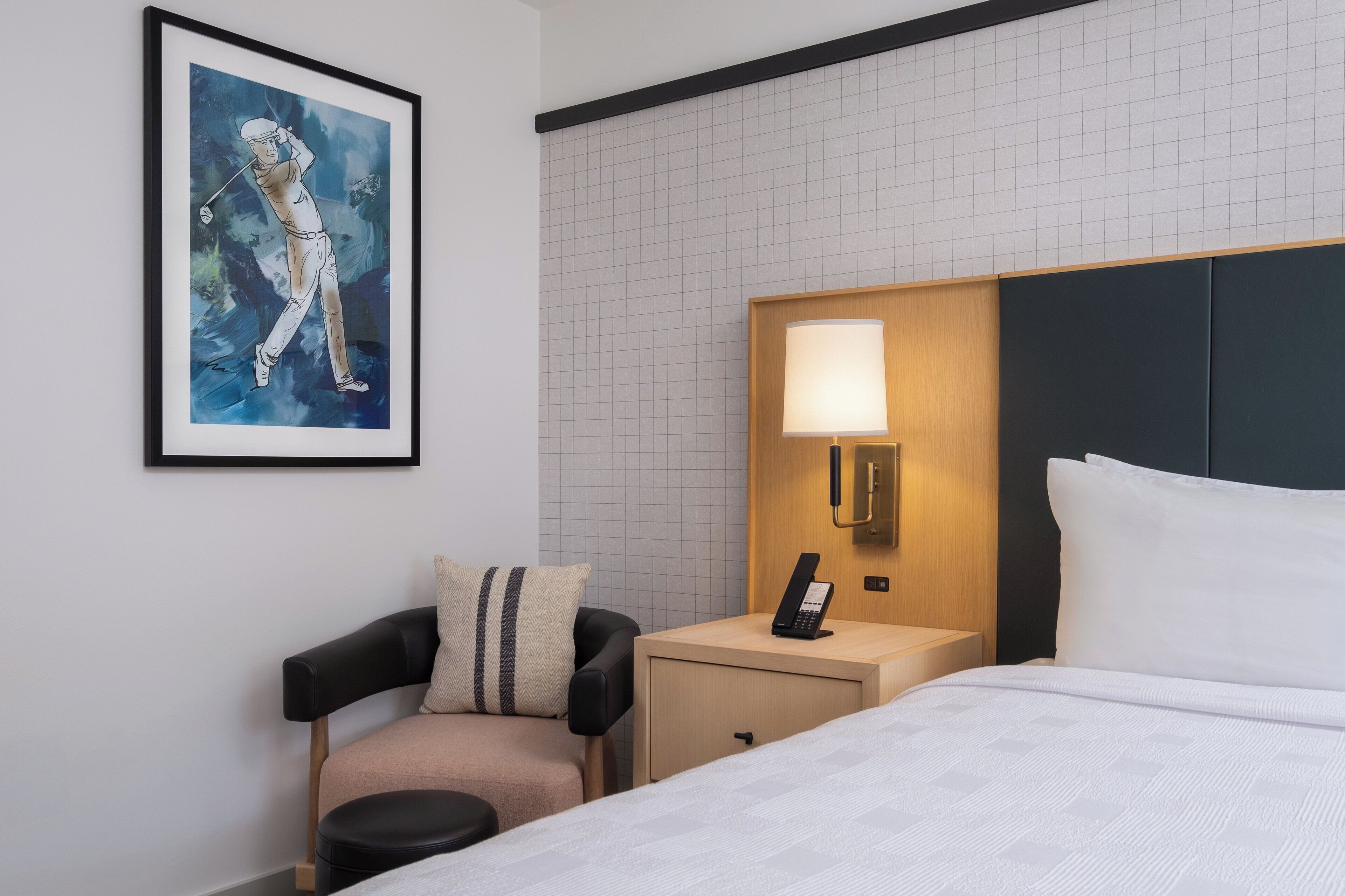 Quarto de hotel com uma cama, uma escrivaninha com luminária, telefone, cadeira e uma obra de arte emoldurada na parede.