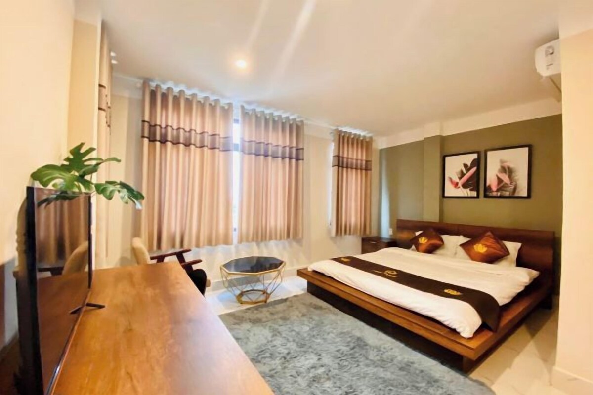 Quarto de hotel com cama, escrivaninha, cadeira, mesa pequena e planta.