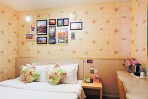 Quarto de hotel com cama, papel de parede floral, mesa de cabeceira com telefone e um vaso de flores, e uma parede com quadros.
