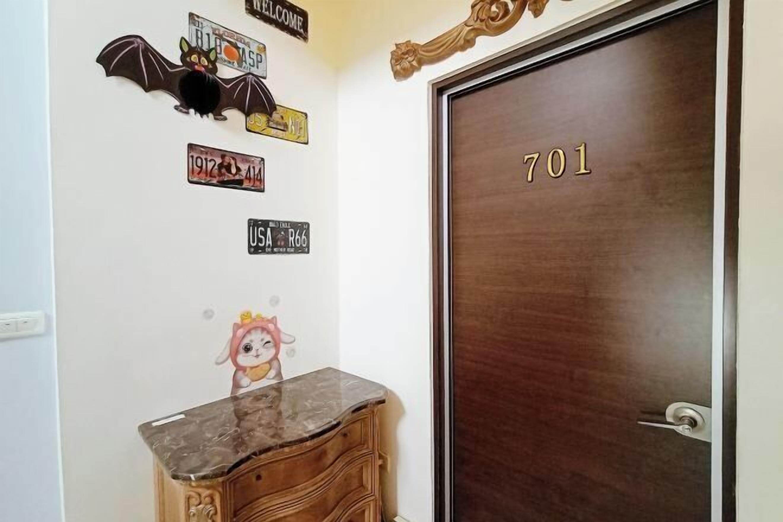 Quarto com porta de madeira identificada com '701', penteadeira com tampo de mármore e decoração nas paredes incluindo um morcego, uma placa de boas-vindas, uma placa de carro e um personagem de desenho animado.