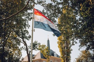 Uma bandeira hasteada em um mastro, com um edifício ao fundo.