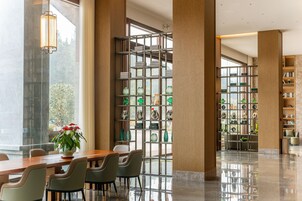 Um hall amplo com balcão de atendimento em madeira, assentos confortáveis, uma planta decorativa e um painel de vidro com prateleiras de objetos decorativos.