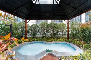 Piscina com borda azulejada, rodeada por vegetação e uma área coberta com telhado de metal.