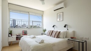 Quarto moderno com uma cama grande, mesas de cabeceira, janela com vista para a cidade e escrivaninha com uma planta e livros.