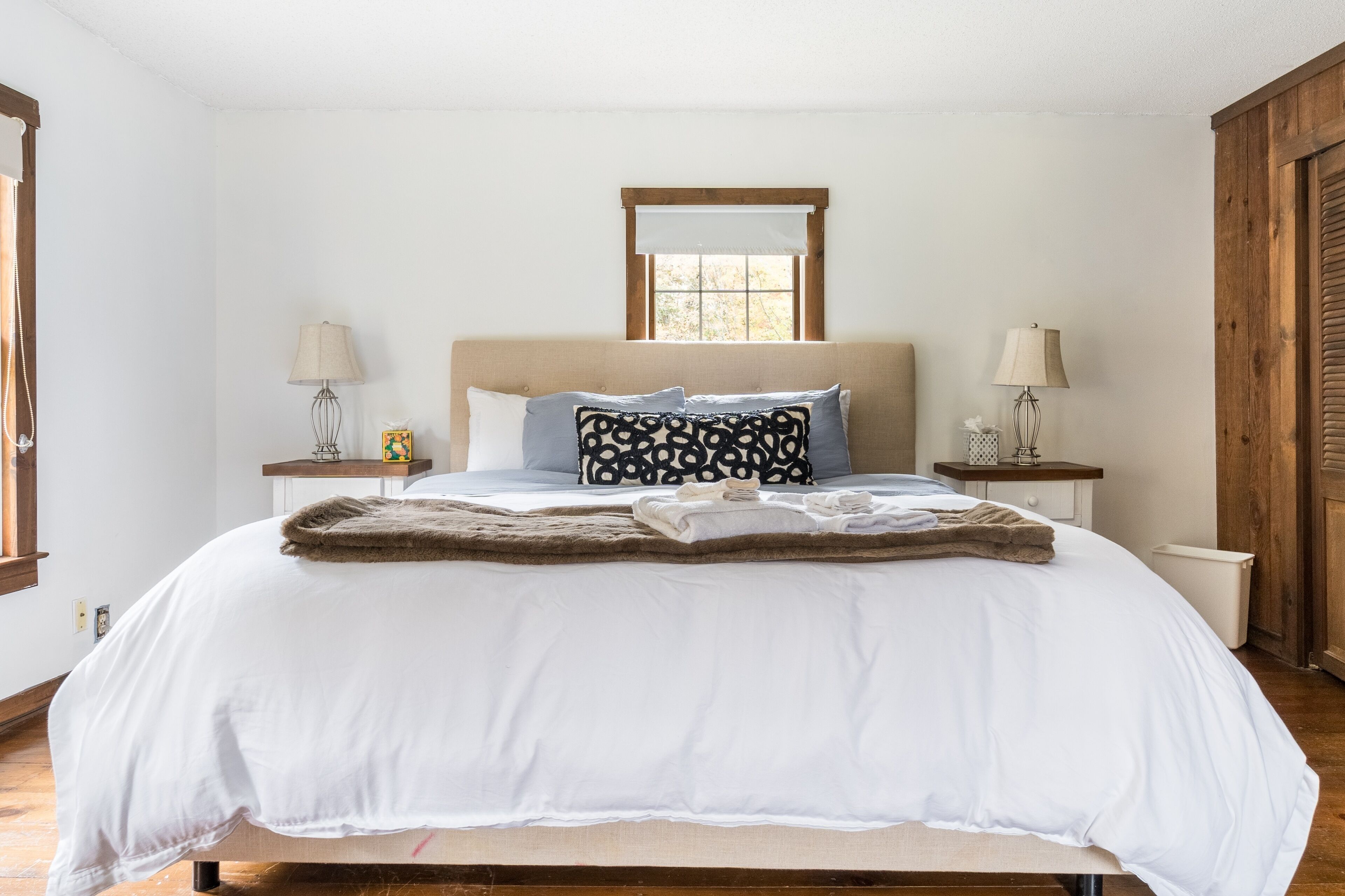 Cama bem arrumada com edredom branco, duas almofadas e um cobertor. Duas mesinhas de cabeceira com luminárias e um objeto decorativo em uma delas. Uma janela com moldura de madeira e cortinas brancas está acima da cama.