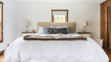 Cama bem arrumada com edredom branco, duas almofadas e um cobertor. Duas mesinhas de cabeceira com luminárias e um objeto decorativo em uma delas. Uma janela com moldura de madeira e cortinas brancas está acima da cama.