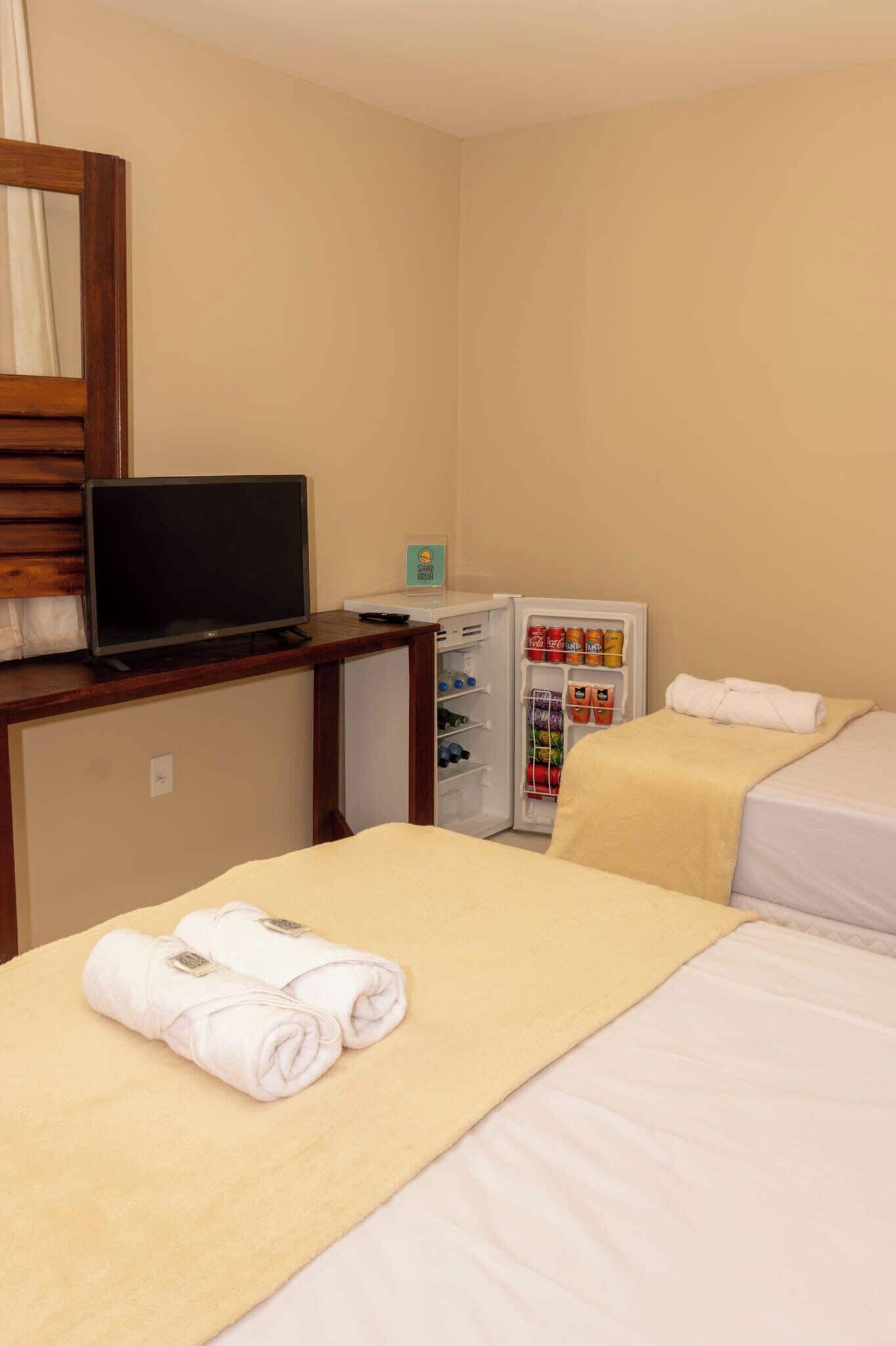 Quarto de hotel com duas camas, uma televisão sobre uma escrivaninha de madeira, um mini-frigorífico e toalhas nas camas.