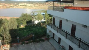Um edifício de vários andares com varanda, um pátio com árvores e vista para um lago e colinas.