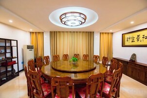 Uma sala de conferências com uma mesa redonda, cadeiras de madeira, luminária no teto, estante de livros e um ar-condicionado fixado na parede.