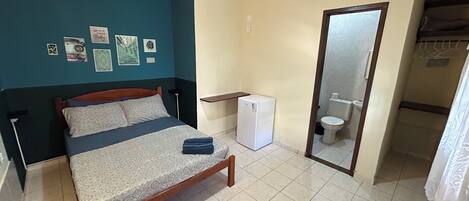 Quarto com cama, um pequeno frigorífico branco, sanita e espelho.