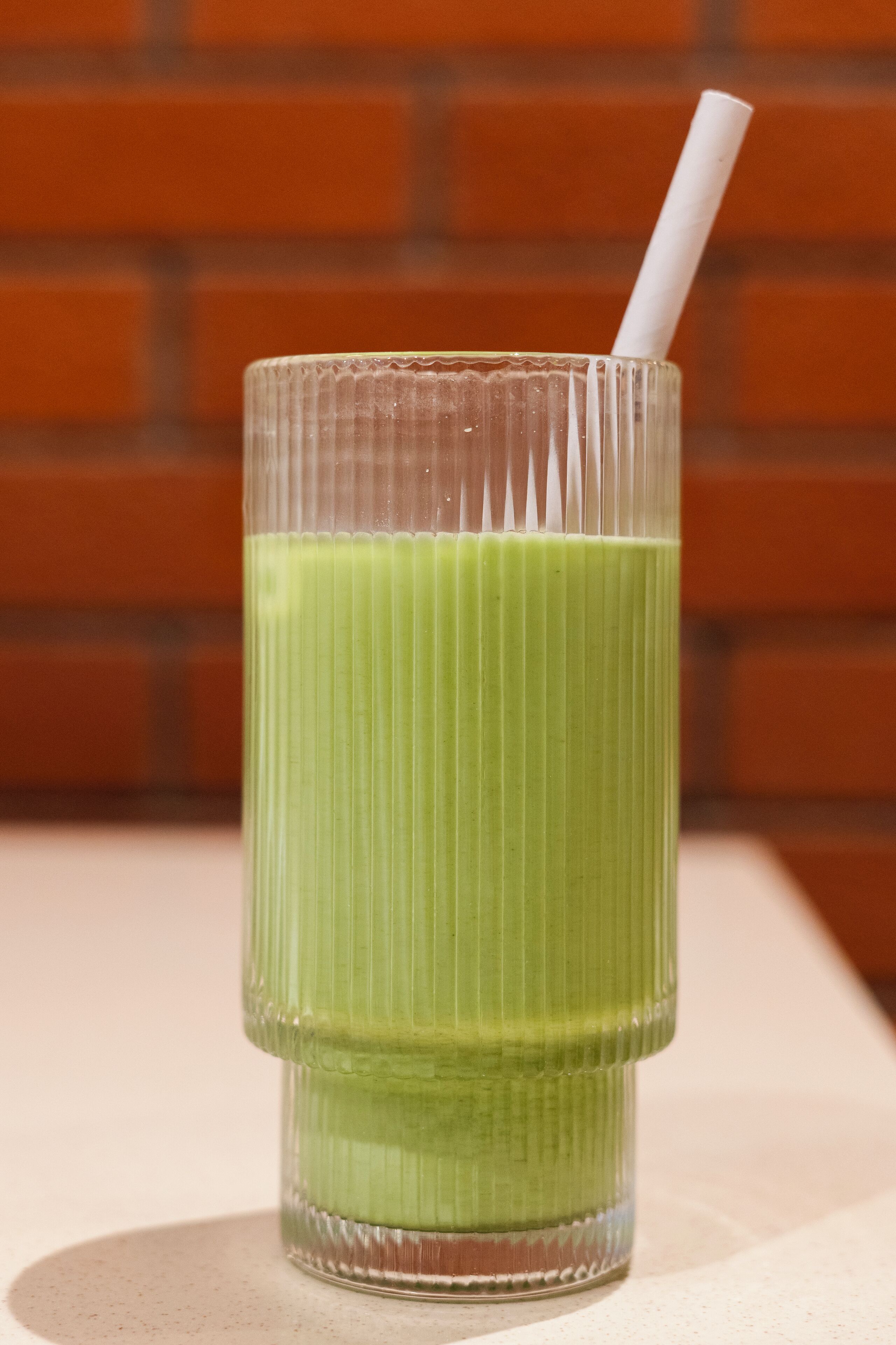 Um copo com canudo cheio de líquido verde, apoiado em uma superfície com parede de tijolos ao fundo.