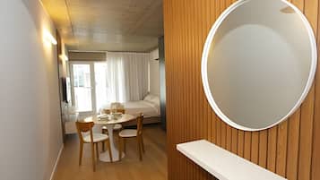 Quarto de hotel com um espelho redondo, painéis de madeira, uma cama, uma mesa pequena com cadeiras e vista para o exterior.