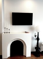 Um quarto moderno com lareira branca, uma televisão de tela plana acima dela e um abajur preto ao lado da lareira.