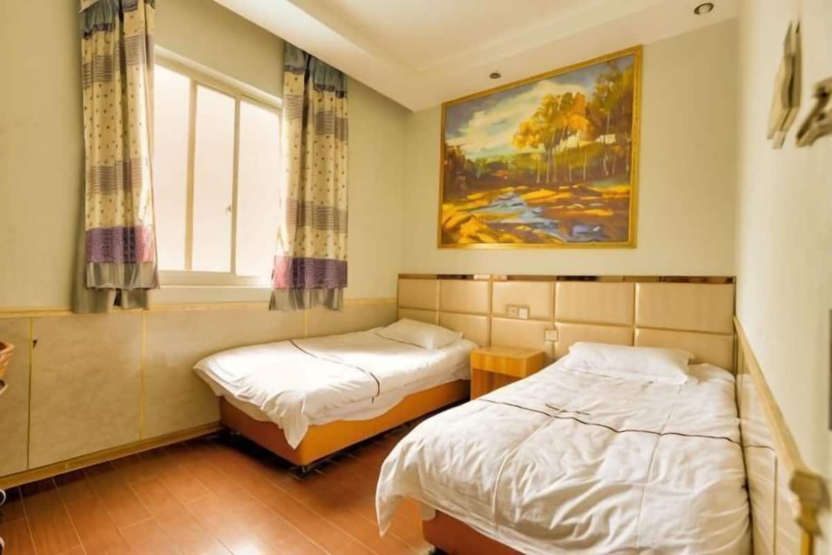Quarto com duas camas, janela com cortinas, um quadro na parede e piso de madeira.