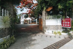 Entrada com portão, placa indicativa, acesso por alameda pavimentada e um edifício com telhado vermelho ao fundo.