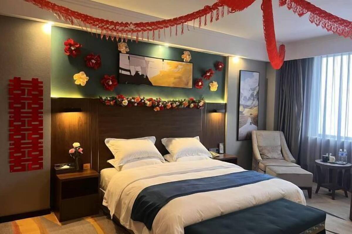 Quarto de hotel com cama, uma cadeira, uma mesinha e uma parede decorada com flores e um quadro.