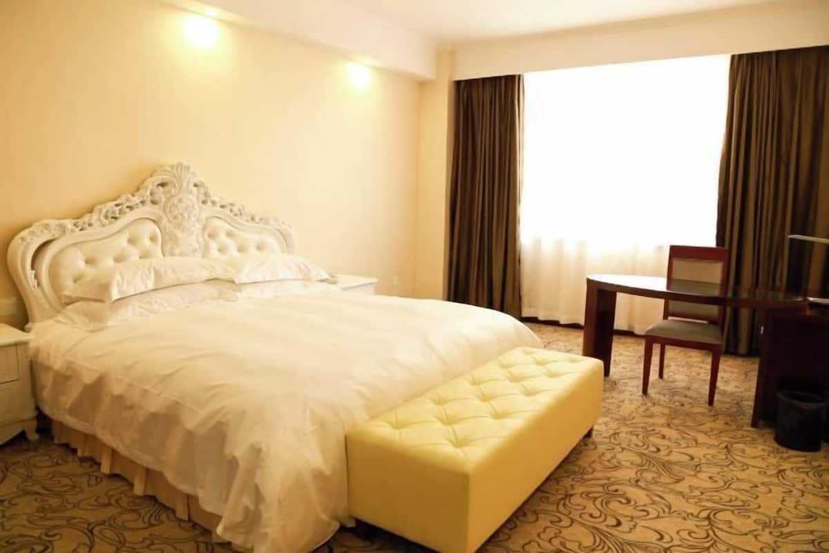 Quarto de hotel com uma cama grande, uma mesinha, uma cadeira e uma janela com cortinas.