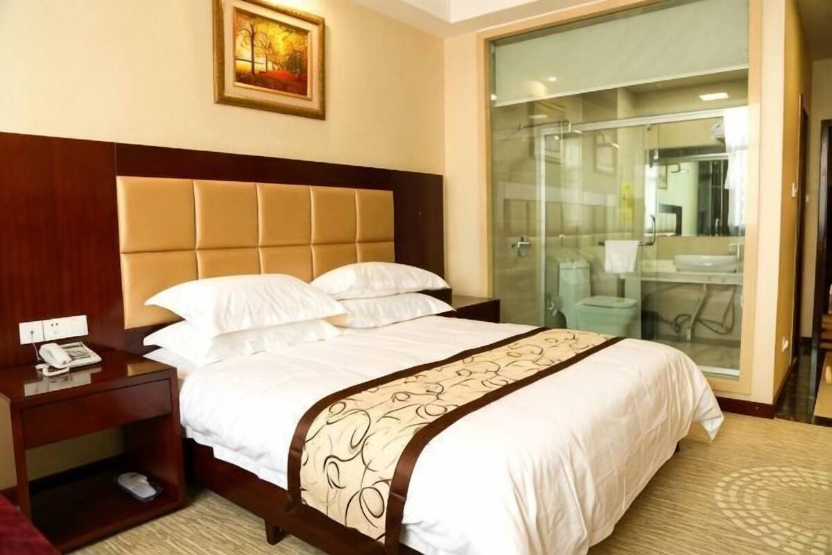 Quarto de hotel com cama, mesa de cabeceira e área de chuveiro com divisória de vidro.